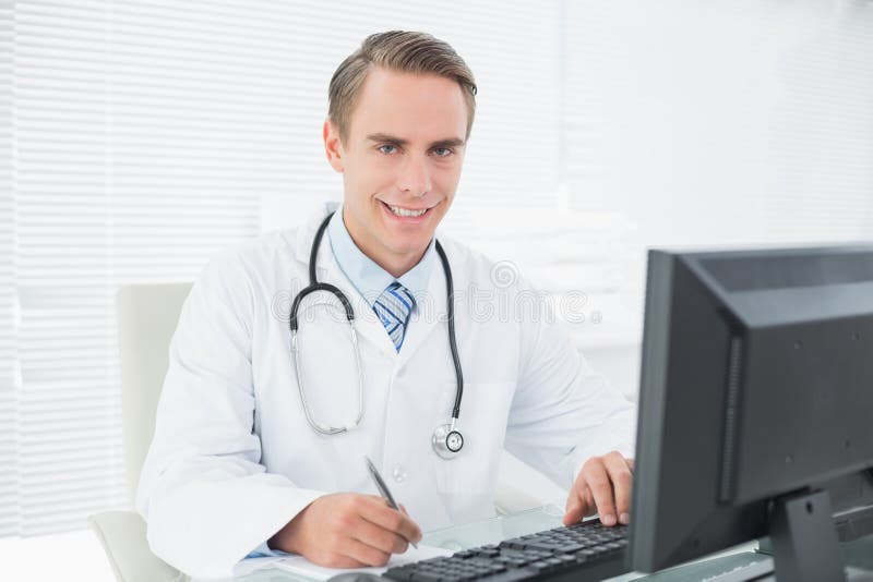 Smiling Doctor Writing Note while Using Computer at Medical Office ...