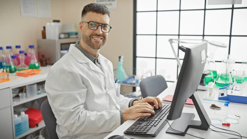 A Smiling Handsome Hispanic Man in a Lab Coat Typing at a Computer in a ...