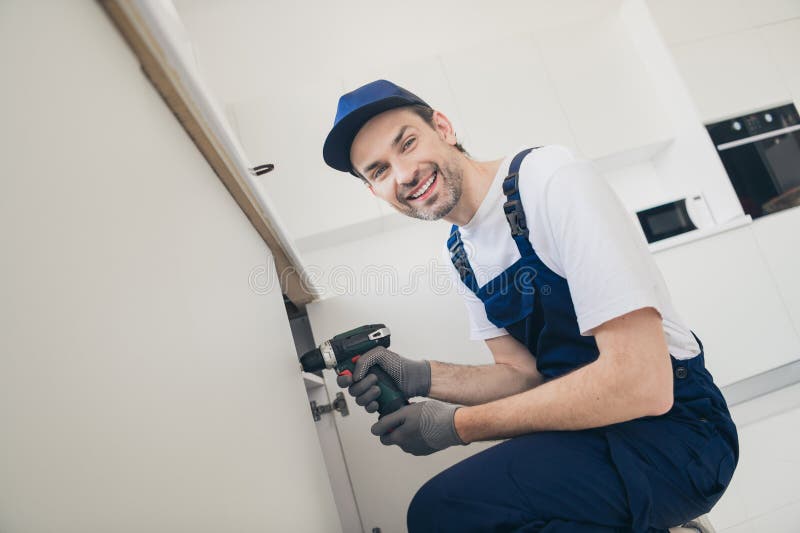 Smiling Handyman in Blue Overalls Using a Cordless Drill for Home Renovation in a Modern Kitchen ...