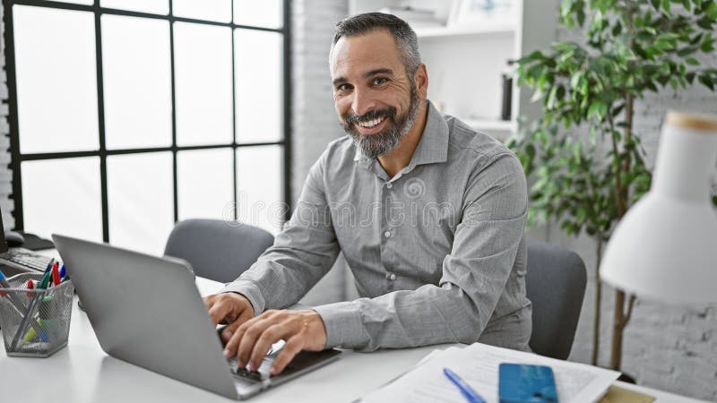 Smiling Hispanic Senior Man with a Grey Beard Working on a Laptop in a ...