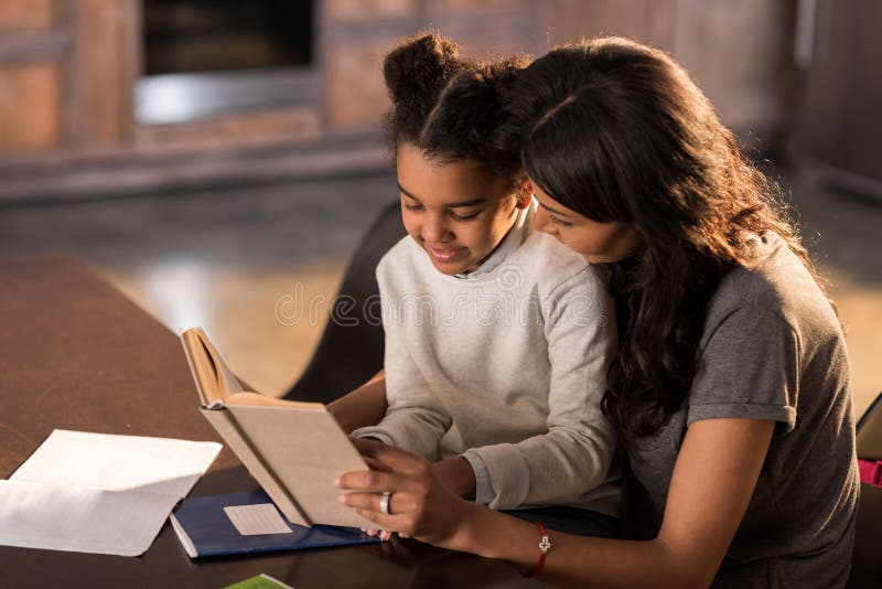 Smiling Mother and Daughter Sitting Together at Table and Reading Book ...