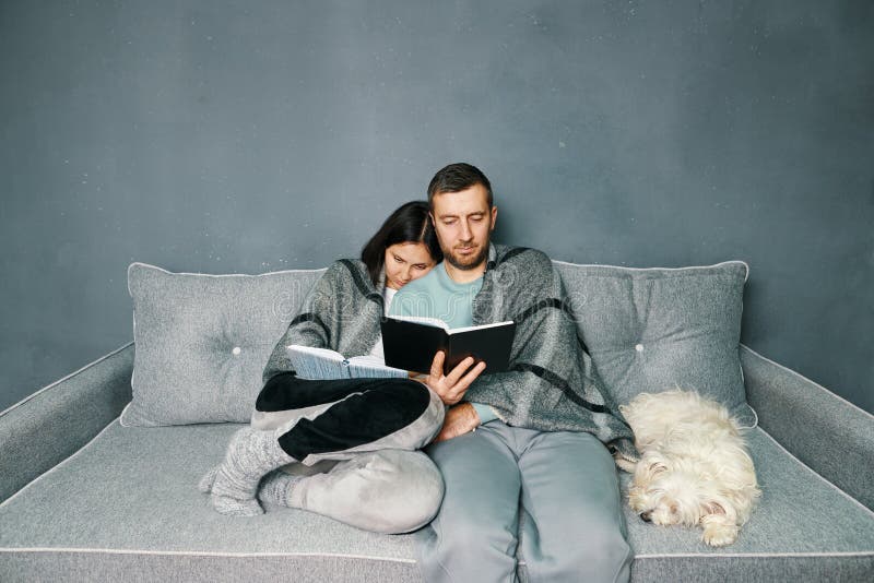 Smiling Young Couple Reading Books Together On Couch Under Plaid At