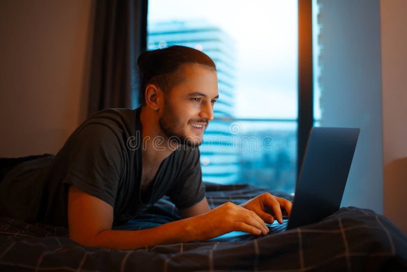 Smiling Young Guy Lying on Bed and Working Home on Laptop. Typing on ...