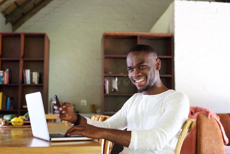 Smiling Young Guy Sitting at Table with Cell Phone and Laptop Stock ...