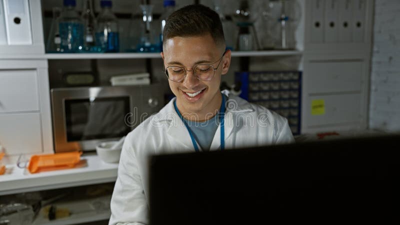 Smiling Young Hispanic Man in Lab Coat Working on Laptop in a ...