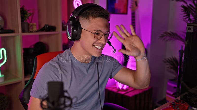 Smiling Young Man with Headphones in a Vibrant Gaming Room Waves at the ...