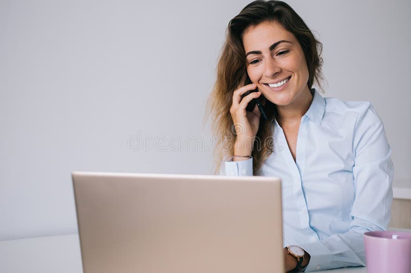 Smiling Young Woman Talking on Smartphone and Typing on Laptop Stock ...