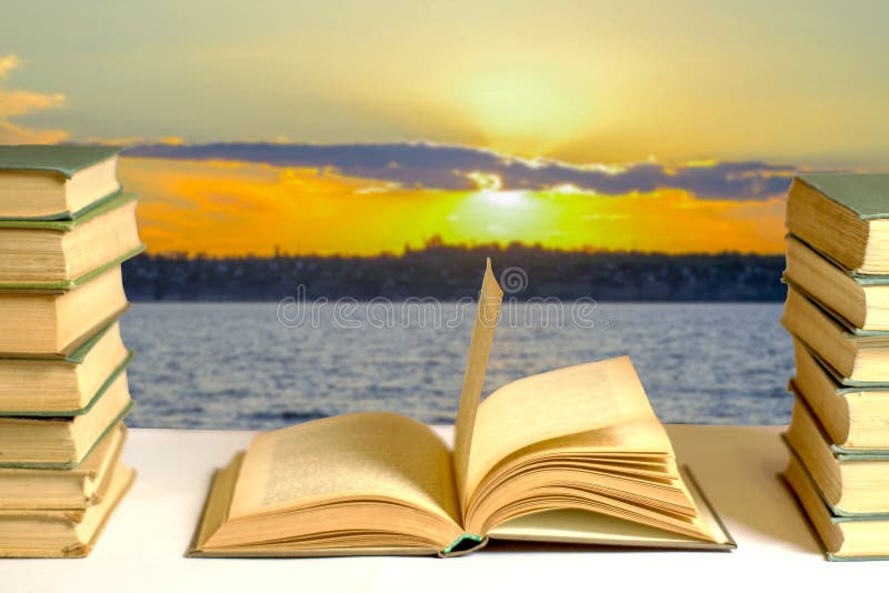 A Stack of Books and an Open Book Against the Backdrop of a Sunset Over ...