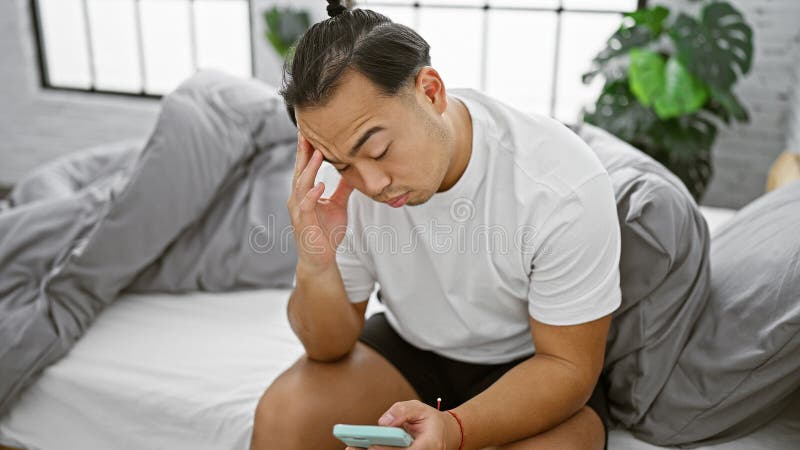 Stressed Young Chinese Man, Sitting in Bedroom, Using Smartphone Stock ...