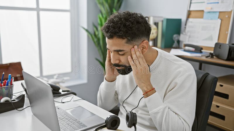 A Stressed Young Hispanic Man with a Beard in a Modern Office, Touching ...