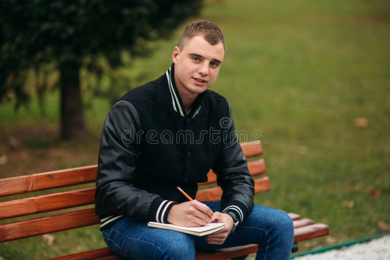 A Student in a Black Jacket Sits in a Park on a Bench Writes Down His ...