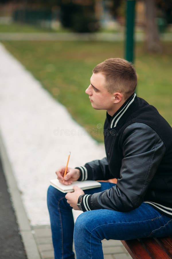 Student in a Black Jacket Sits in a Park on a Bench Writes Down His ...