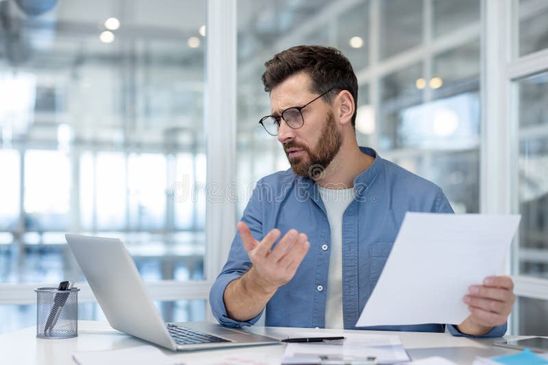 A Thoughtful Businessman in Glasses Analyzes Documents, Puzzled while ...