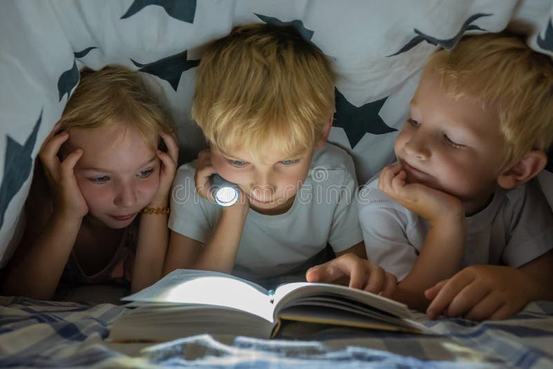 Three Young Children are Reading a Book with a Flashlight Under the ...