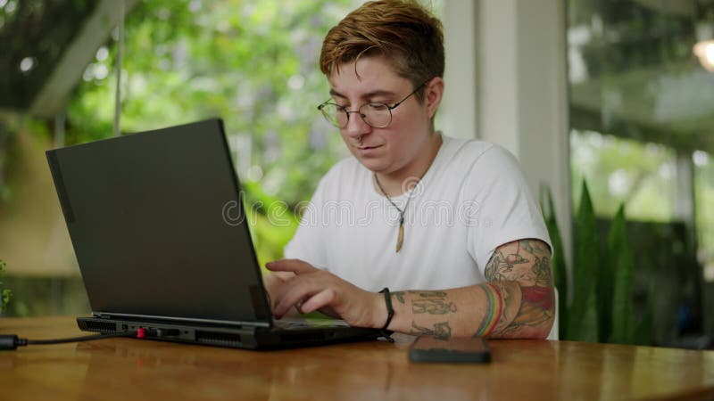 Transgender Individual Works on Laptop in Bright Cafe, Greenery Visible ...