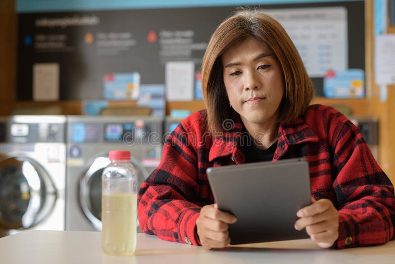 Transgender Non-binary Individual Sitting in a Modern Laundromat Laundry Shop Using Tablet ...