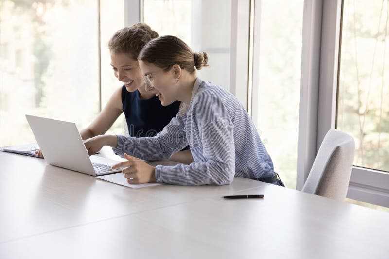 Two Cheerful Young Female Employees Sharing Workplace, Using Online ...