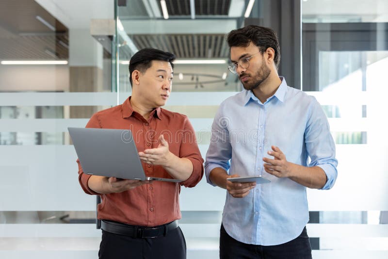 Two Colleagues Discussing Work while Using a Laptop and Tablet in ...