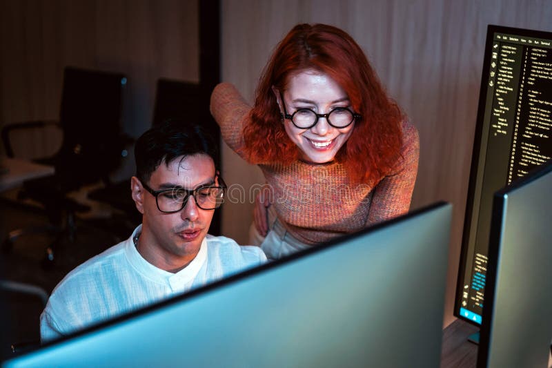 Two Coworkers Collaborate at Dual Monitors in a Dim Office, Reviewing ...