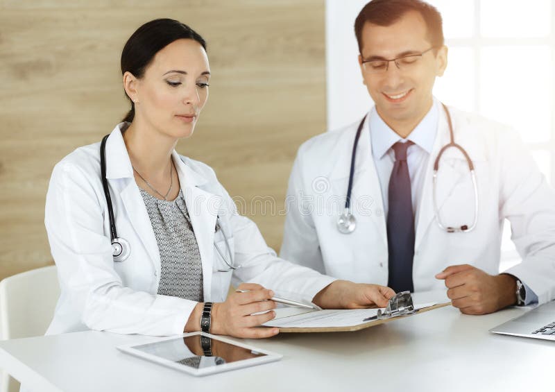 Two Doctors Discussing Treatment Problems while Sitting at the Desk in ...