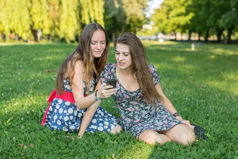 Two Girlfriends Sitting In A Clearing Together Use A Smartphone Stock