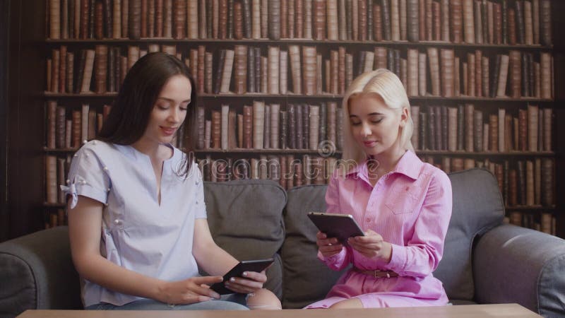 Two Girls Sit in the Library and Read a Book in Electronic Form Stock ...