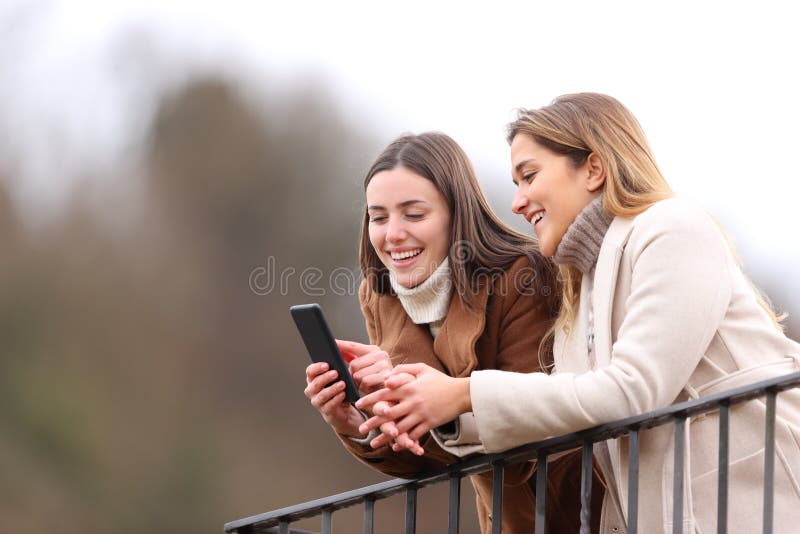 Two Happy Friends Checking Phone in Winter in a Balcony Stock Image ...