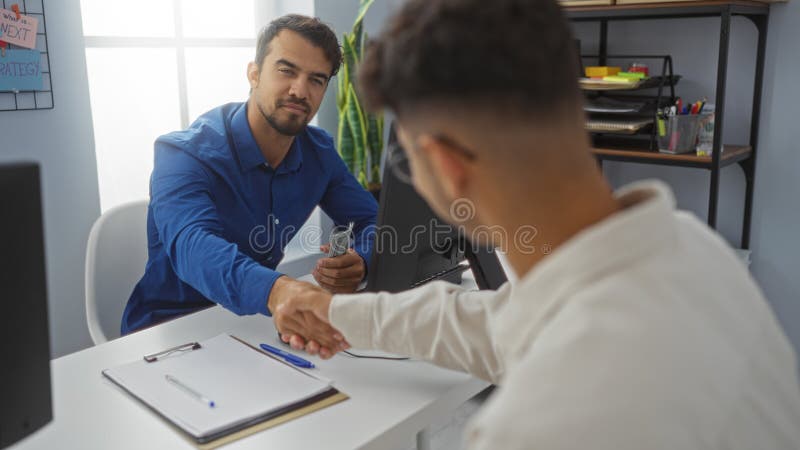 Two Hispanic Men in an Office Handshake Across a Desk Surrounded by ...