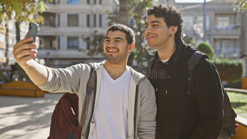 Two Smiling Men Taking a Selfie Together in a Sunlit Urban Park Setting ...