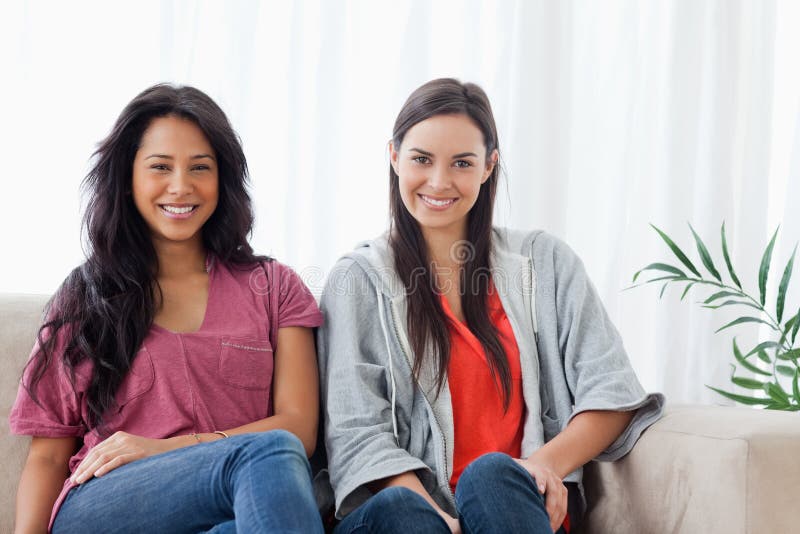 Two Smiling Women Sit On The Couch Together While Looking At The Stock ... Two Smiling Women Sit On The Couch Together While Looking At The Stock ...