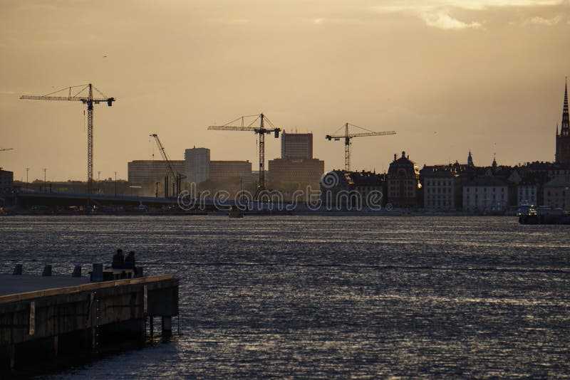 Two Unrecognizable Persons Sit on Pier in Front of City in Sunset Stock ...