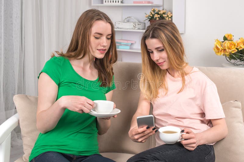 Two Young Attractive Women Sit On A Sofa In The Room And Talk A Stock