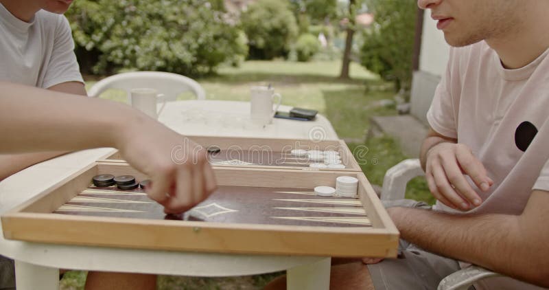 Two Young Men Play a Game of Backgammon at an Outdoor Table Surrounded ...