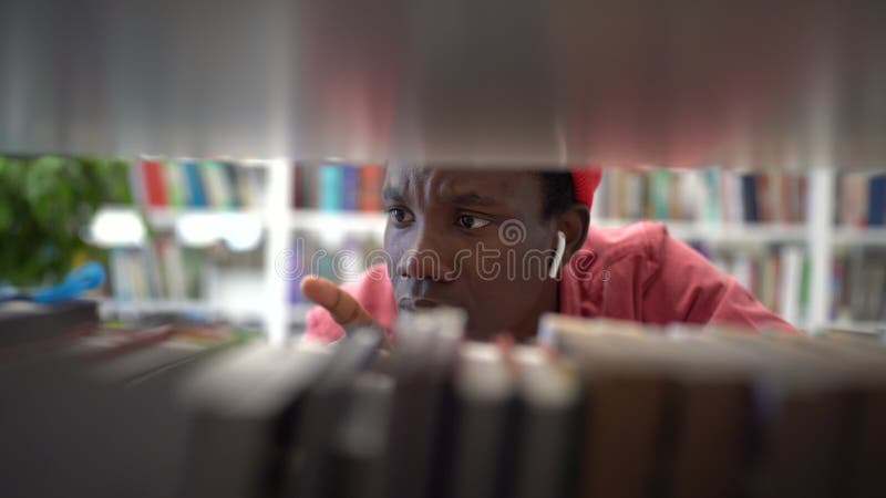 Stressed African Guy Student Looking for Book on Library Shelf, Having Problem Finding Textbook ...