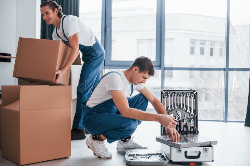 Uses Tools from Case. Two Young Movers in Blue Uniform Working Indoors ...