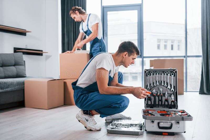 Uses Tools from Case. Two Young Movers in Blue Uniform Working Indoors ...