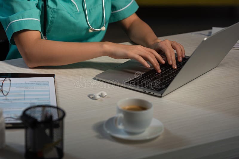 View of Nurse in Uniform Sitting at Table and Using Laptop during Night ...