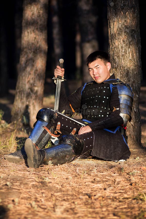 A Warrior in Black Armor with a Sword in His Hands Sits on the Ground ...