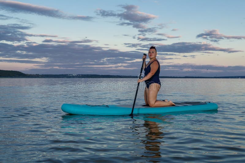 A Woman In A Bathing Suit With A Mohawk Sits On A Sup Board At Sunset