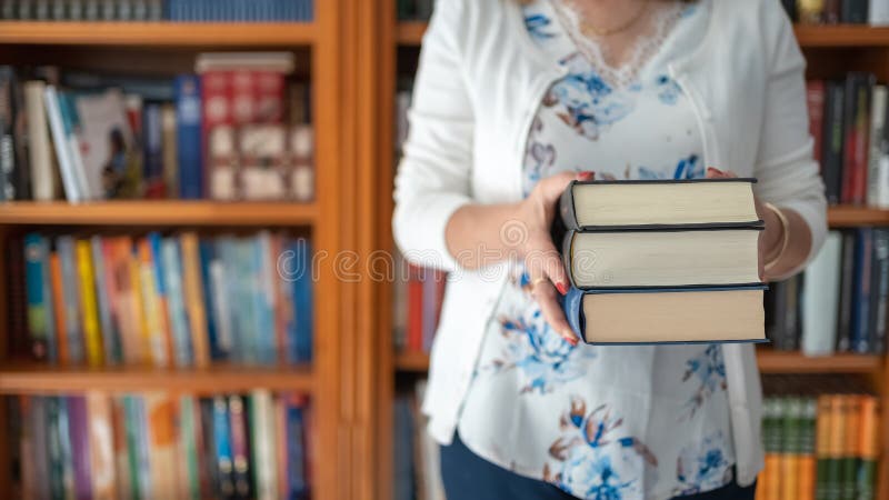 Woman Holding a Stack of Large Books with a Large Bookcase Full of ...