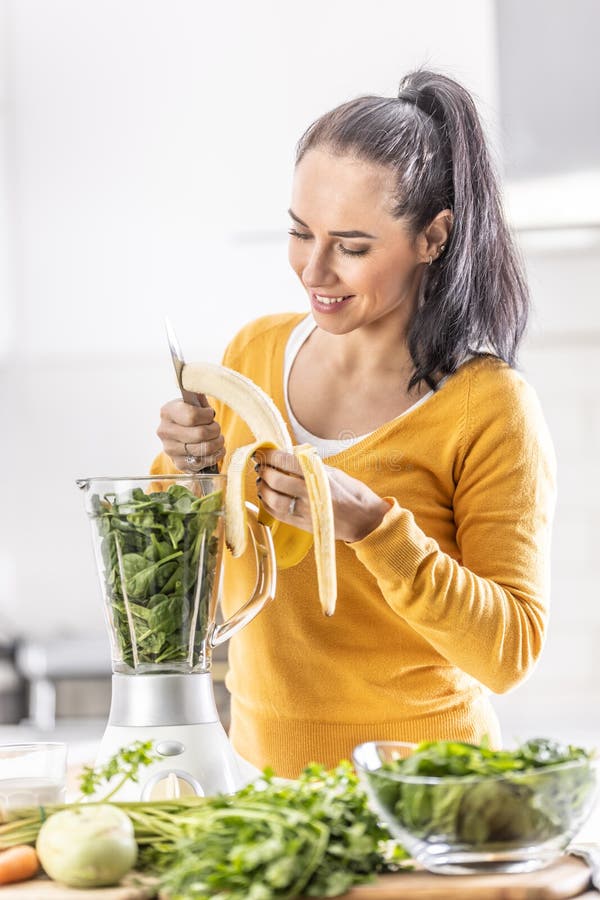 A Woman In The Kitchen Cuts A Banana In A Blender Together With Spinach