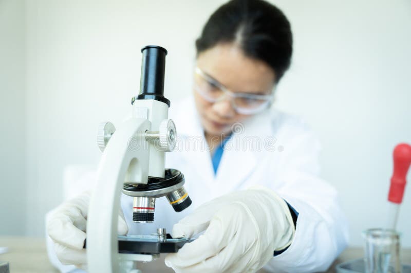 A Woman in a Lab Coat is Looking through a Microscope. she is Wearing ...