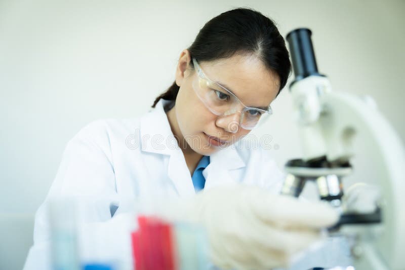 A Woman in a Lab Coat is Looking through a Microscope. she is Wearing ...