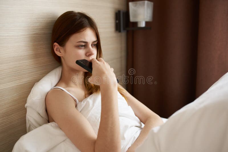Woman Lies In Bed Under The Covers With A Mobile Phone In Her Hand