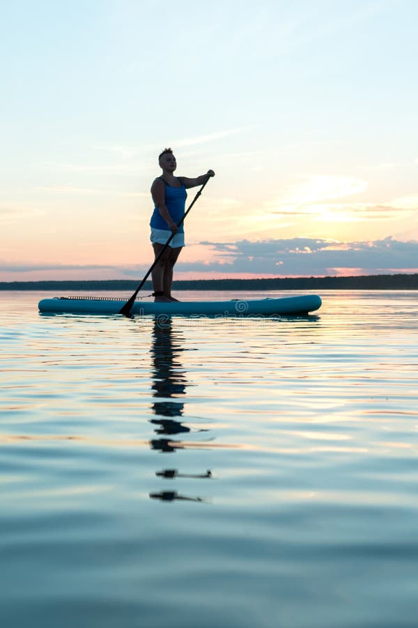 A Man In Shorts Stands On A Sup Board In The Evening At Sunset In The