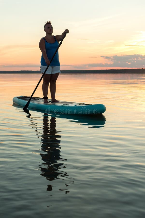 A Man In Shorts Stands On A Sup Board In The Evening At Sunset In The