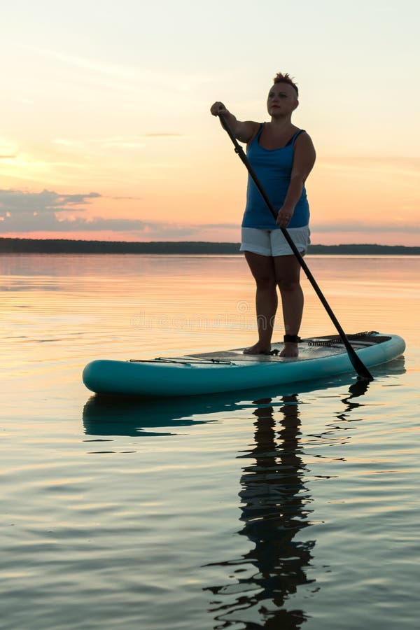 A Woman In Mohawk Shorts Stands On A Sup Board With A Paddle At Sunset