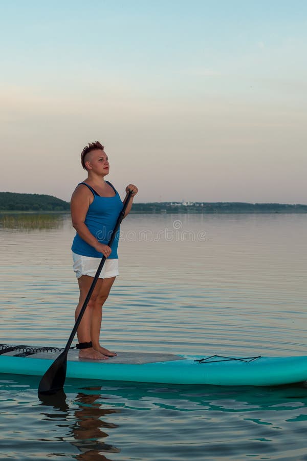 A Man In Shorts Stands On A Sup Board In The Evening At Sunset In The