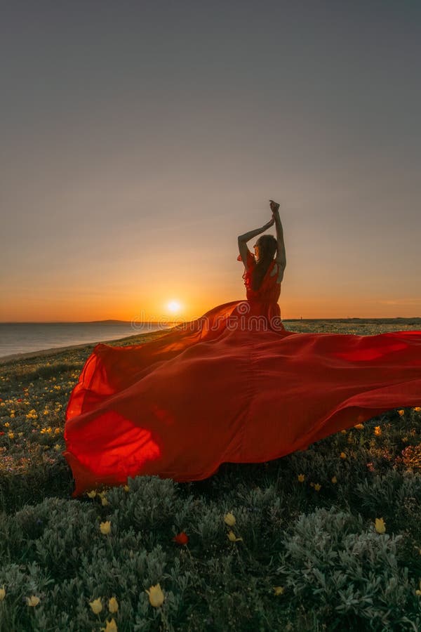 A Woman In A Red Dress Is Standing In A Field With The Sun Setting