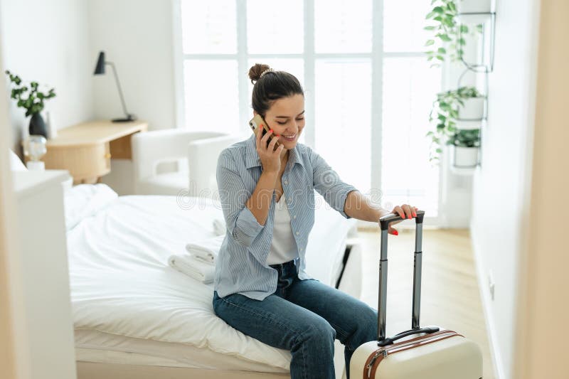 Woman With A Suitcase Sitting On Bed In Hotel Room And Using Phone